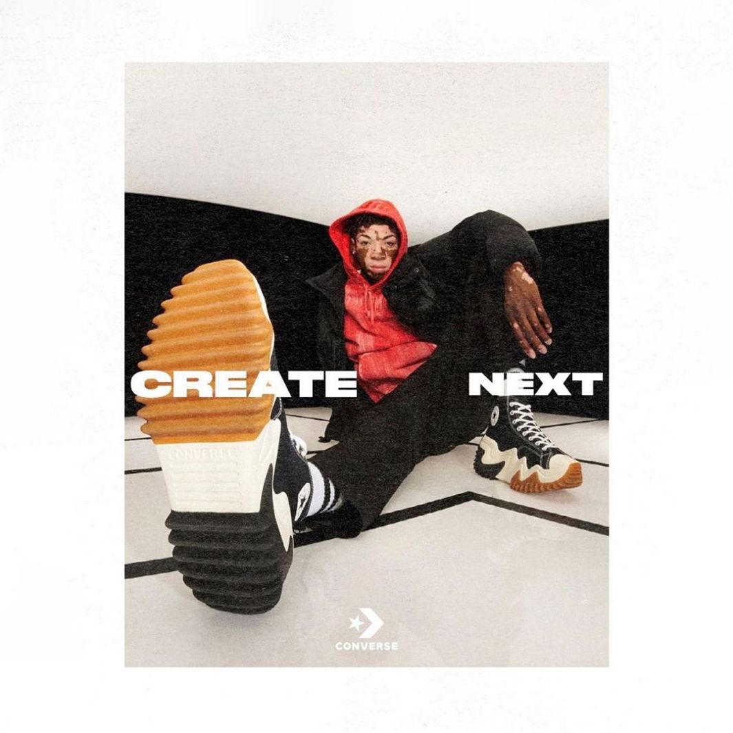 Man sitting on floor bottom of converse shoe facing camera. Text reads Create Next